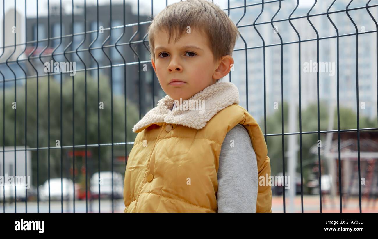 Upset, sad and lonely boy leaning on metal fence at public playground ...