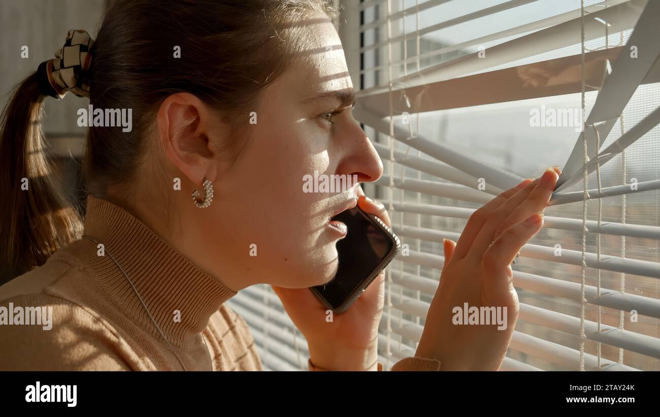 Portrait of stressed and scared woman loooking through window blinds on ...