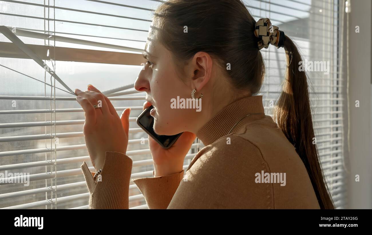 Young scared woman looking through window on street and calling 911 ...