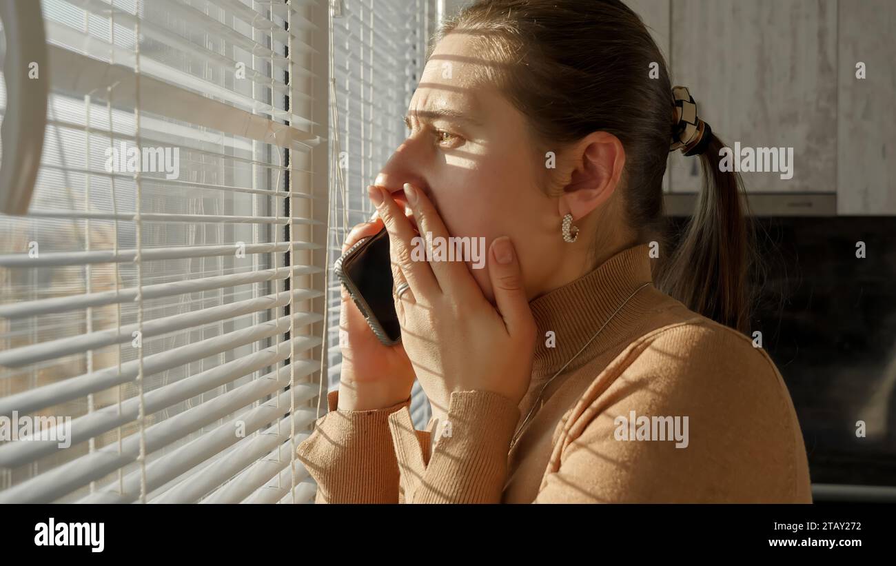 Scared woman talking to 911 emergency service looking outdoors through window blinds. Crime ...
