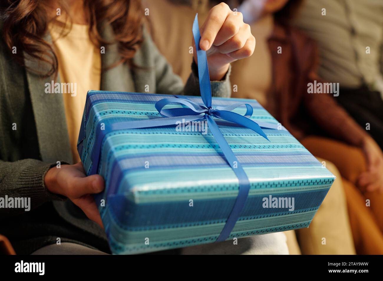 Hand of youthful girl pulling thin blue silk ribbon on top of giftbox ...