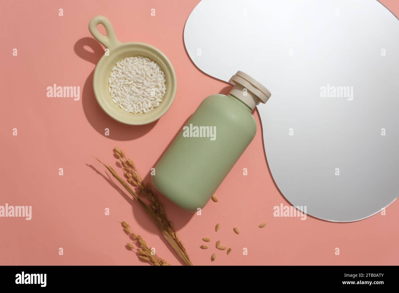 A pastel green cosmetics bottle, bean-shaped mirror, and rice on a pink ...