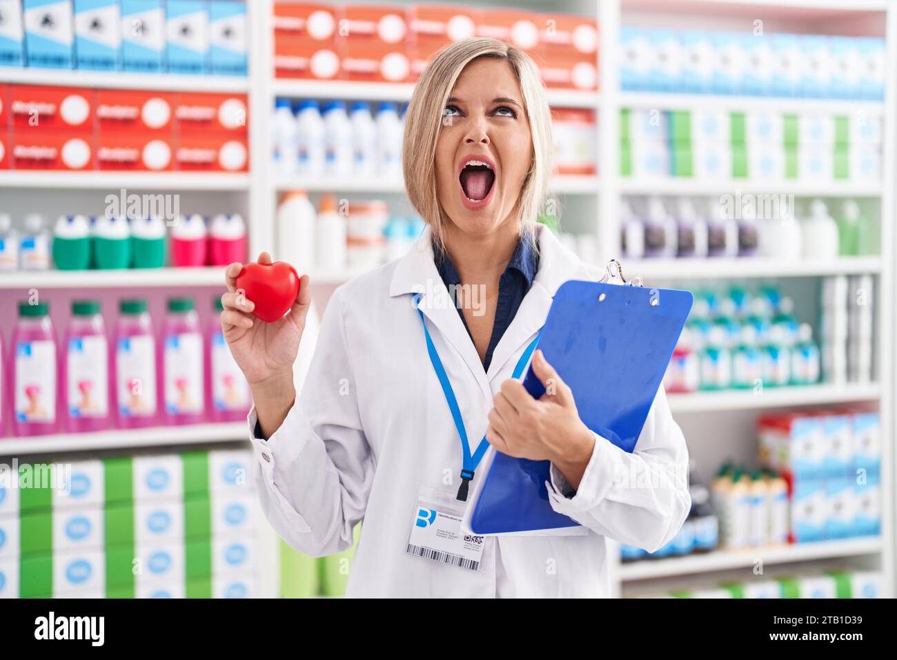 Young woman working at pharmacy drugstore holding heart angry and mad ...