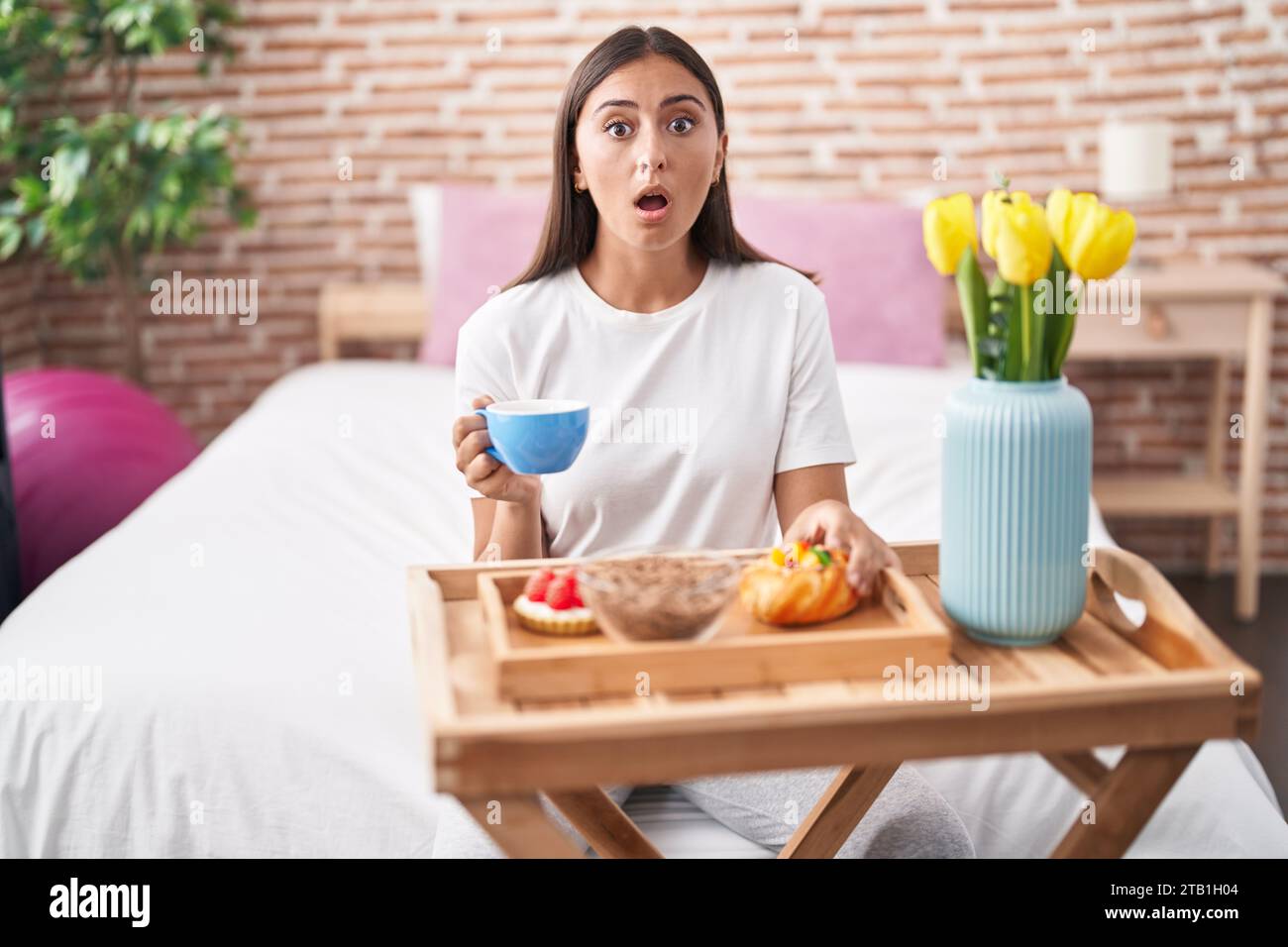 Young hispanic woman eating pastries for breakfast sitting on the bed afraid and shocked with ...