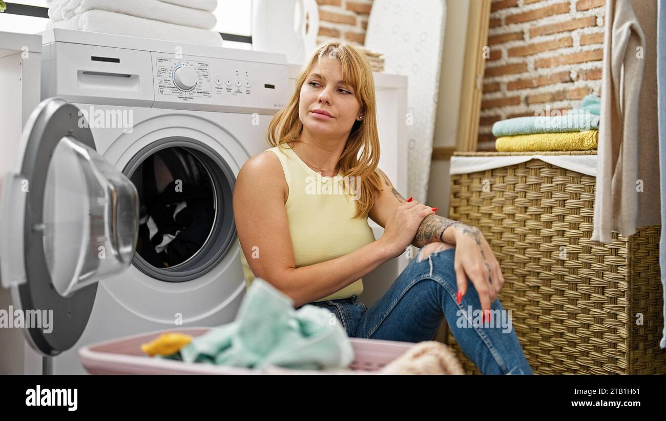 Young blonde woman sitting on floor leaning on washing machine tired at ...