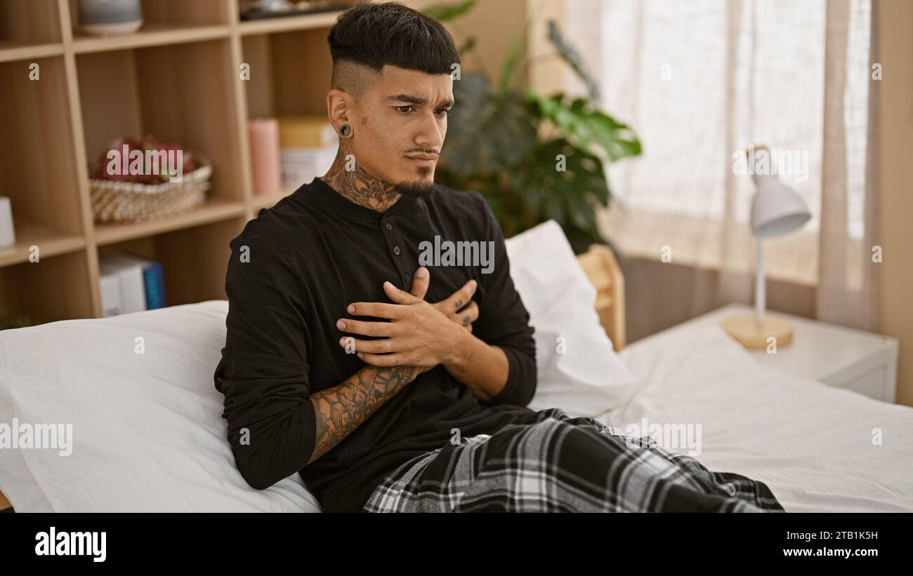 Handsome young latin man, tattooed and suffering, sitting on his bed ...