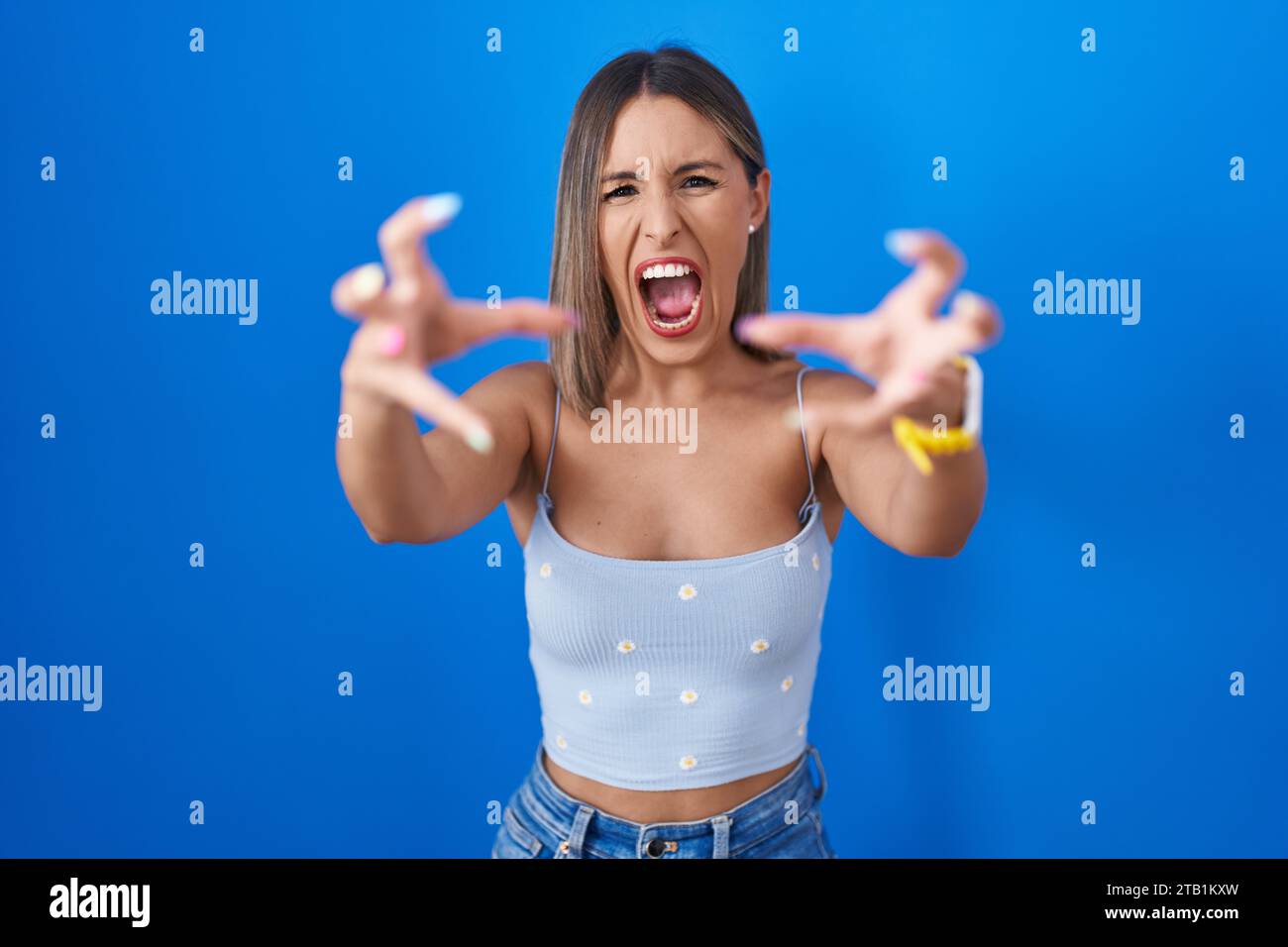 Young woman standing over blue background shouting frustrated with rage ...