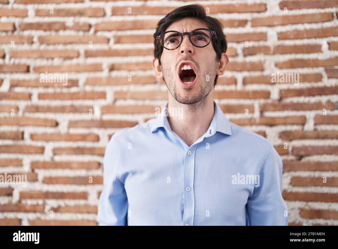 Young hispanic man standing over brick wall background angry and mad ...