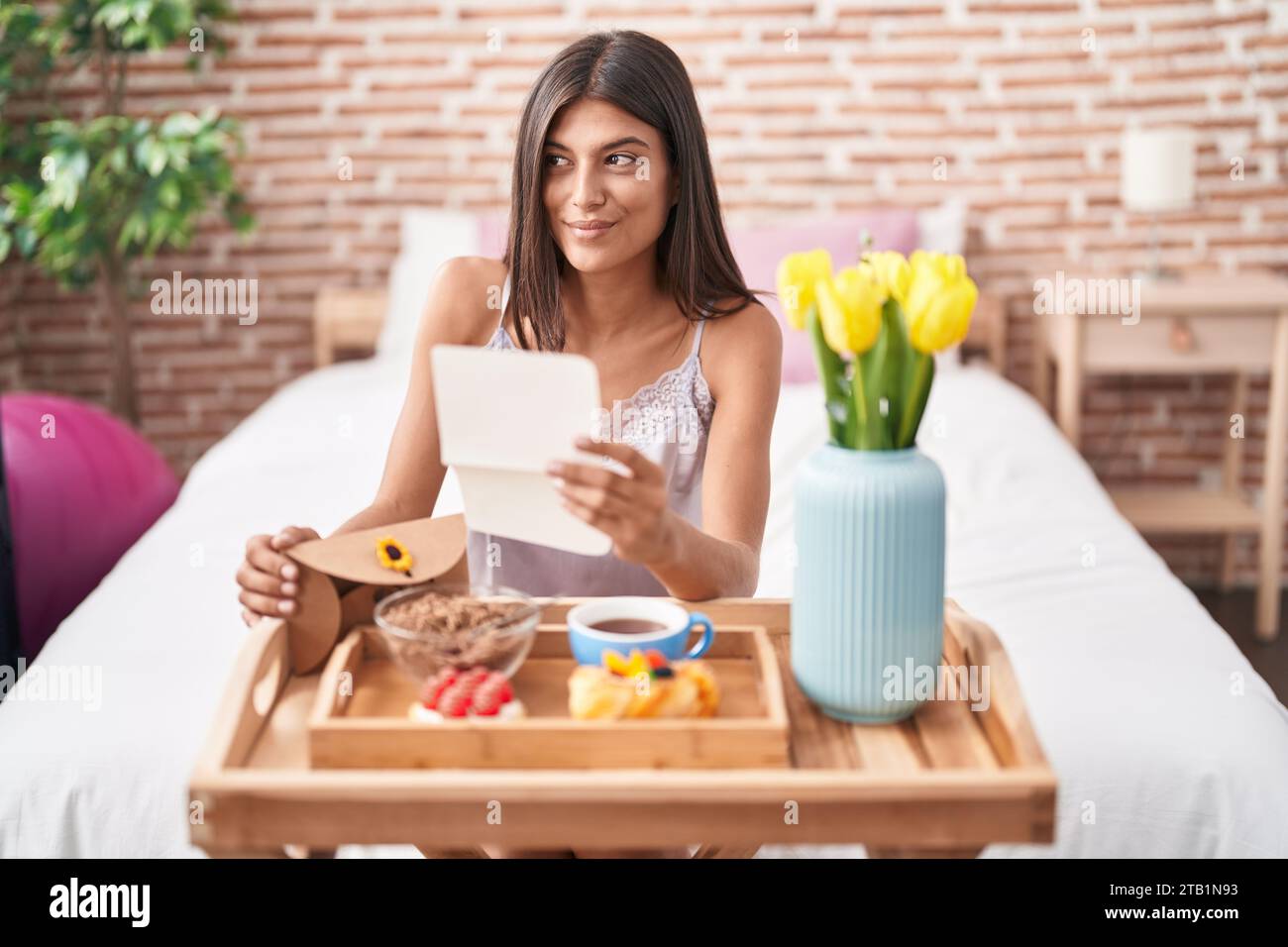 Brunette young woman eating breakfast in the bed reading a letter smiling looking to the side ...