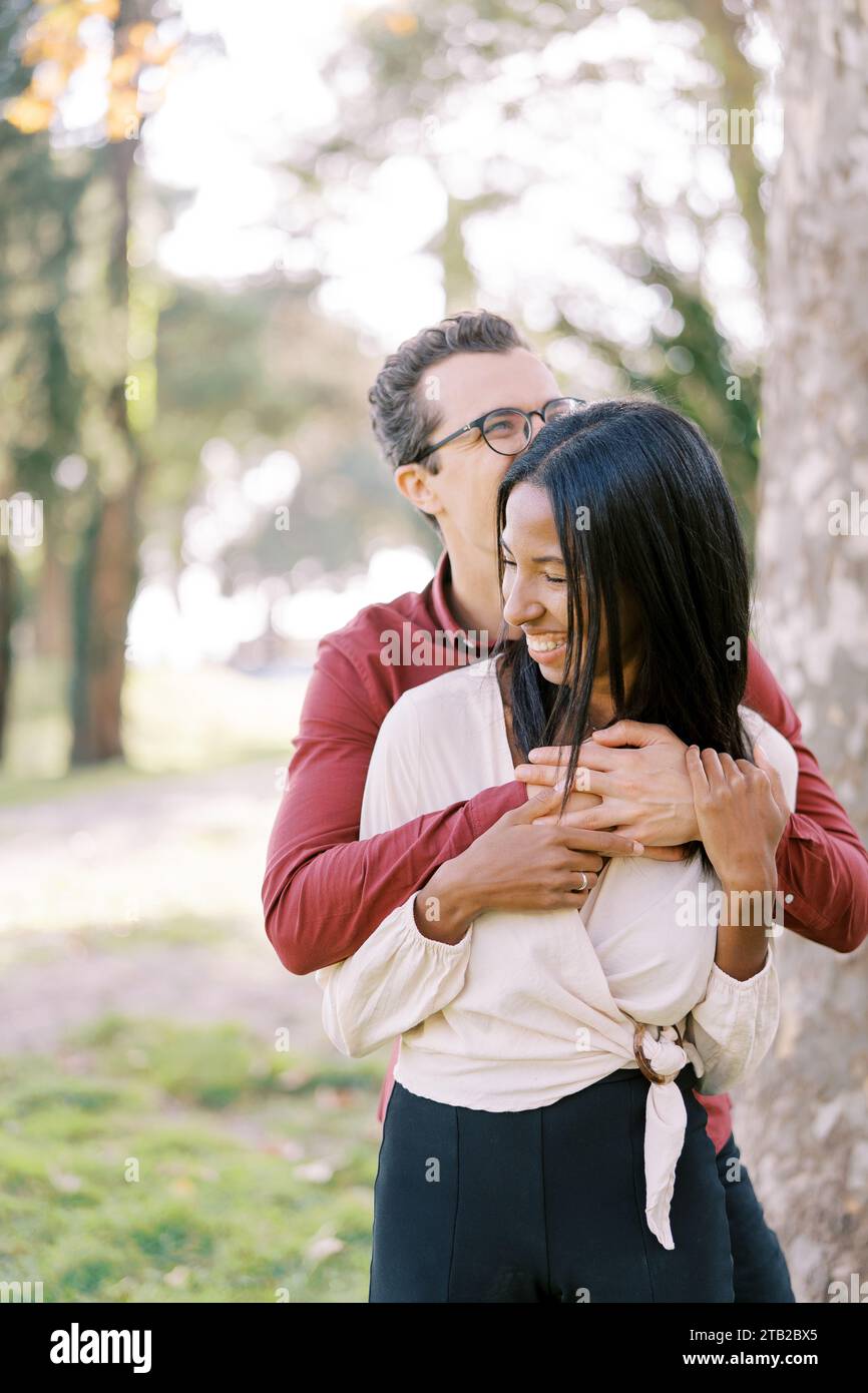Happy man hugs laughing woman from behind by the shoulders and kisses ...