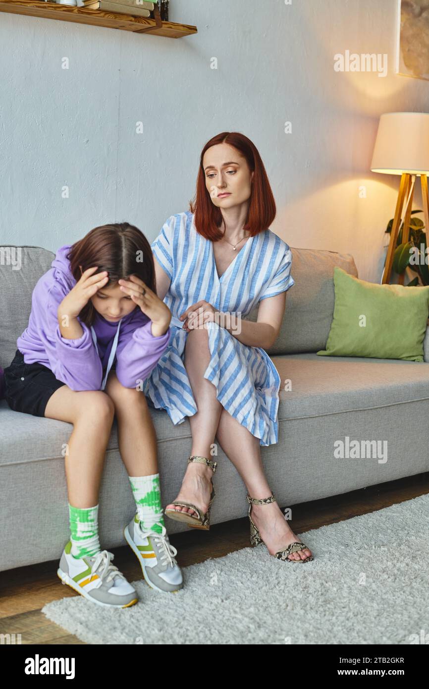 worried mother calming depressed teenage daughter sitting with bowed head on couch at home ...