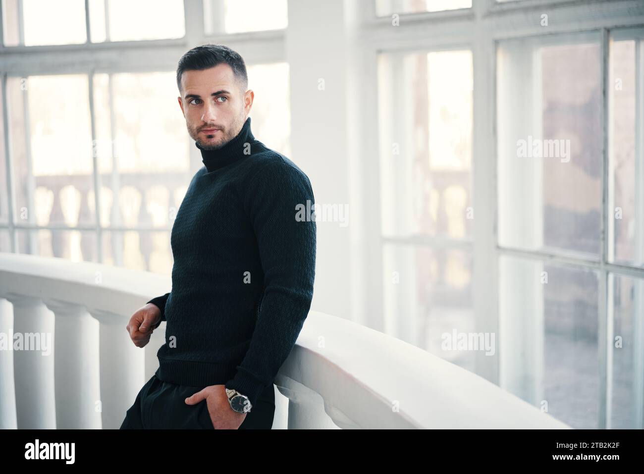 Handsome young man in black turtleneck leans on balcony railings, with ...