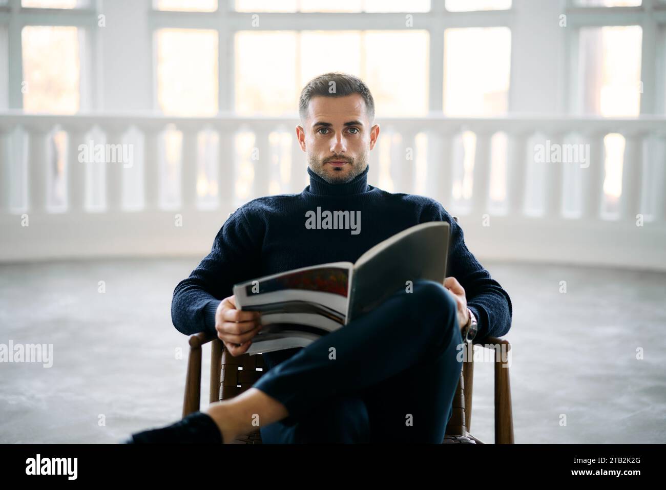 Young handsome man reading magazine seated comfortably in chair in ...