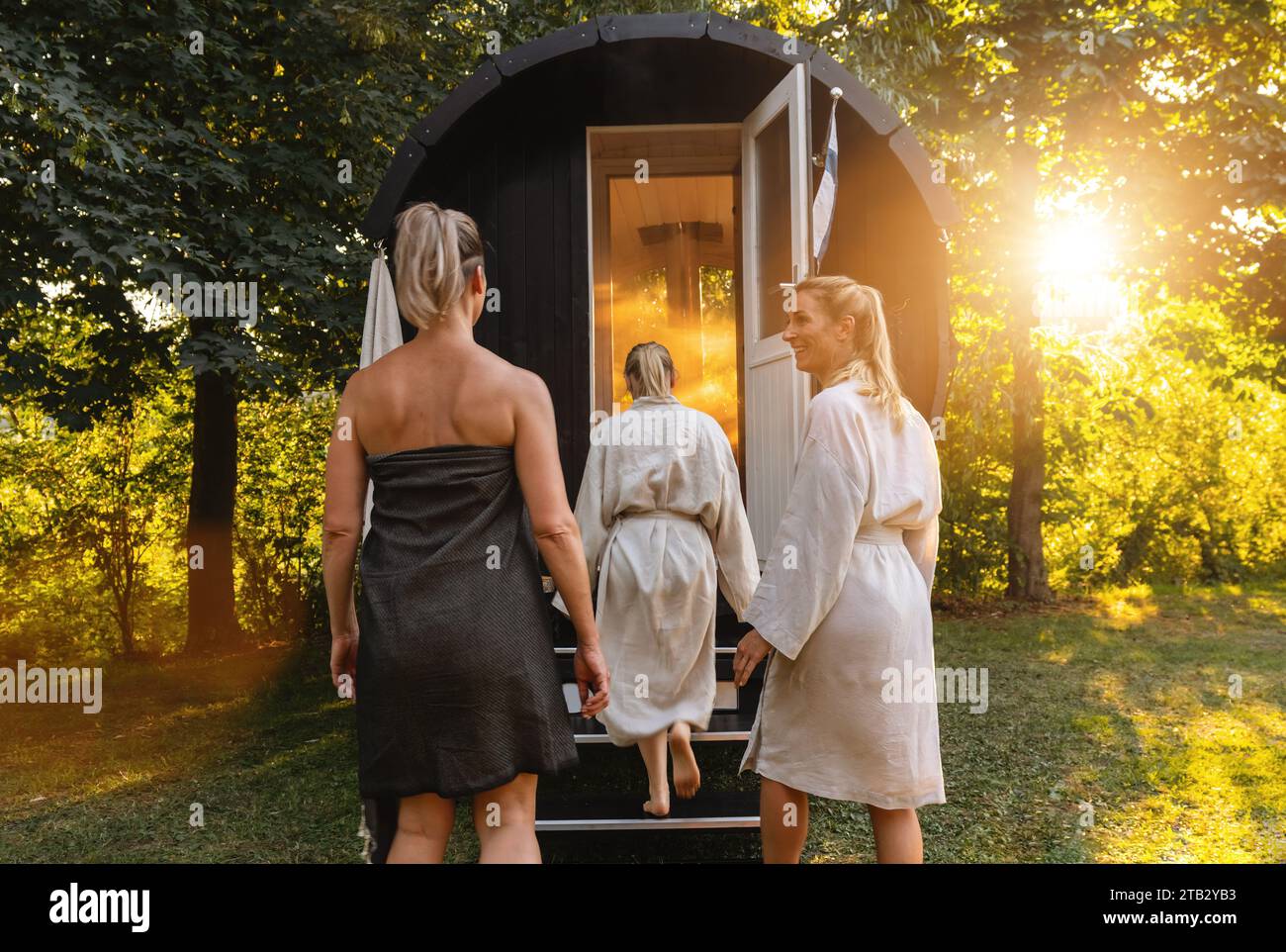 Three people in robes goes in a Finnish sauna barrel in a sunlit forest ...