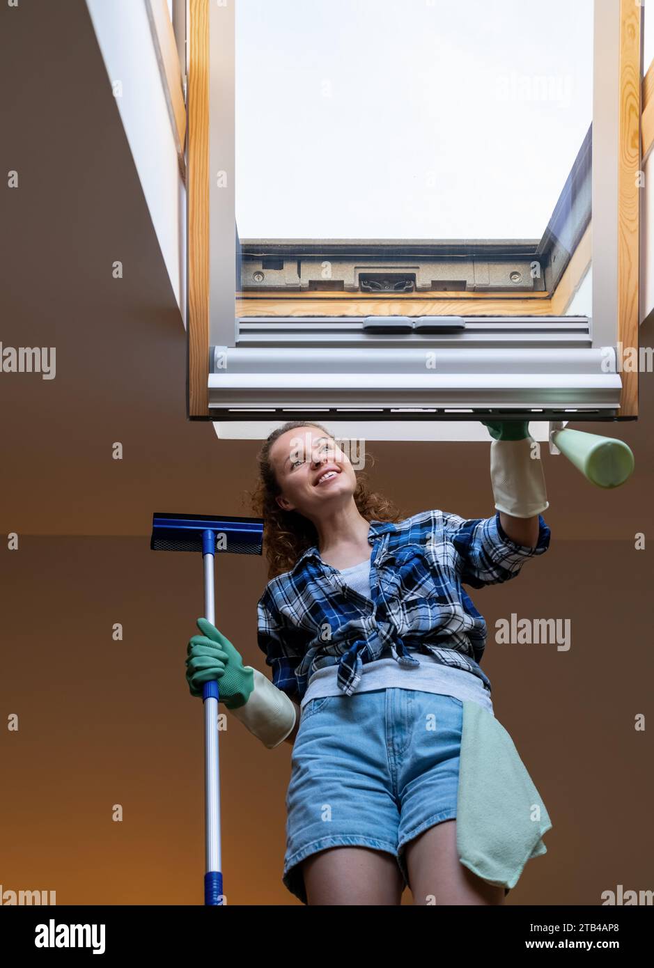 Cheerful young woman cleans skylight window in her apartment. Housework ...