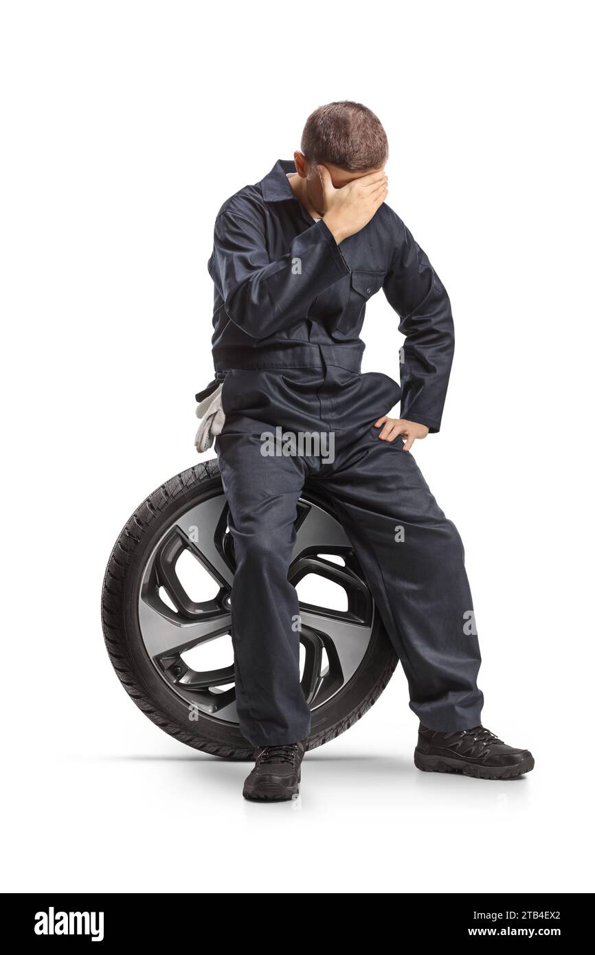Car mechanic in a uniform sitting on a tire and holding his head ...
