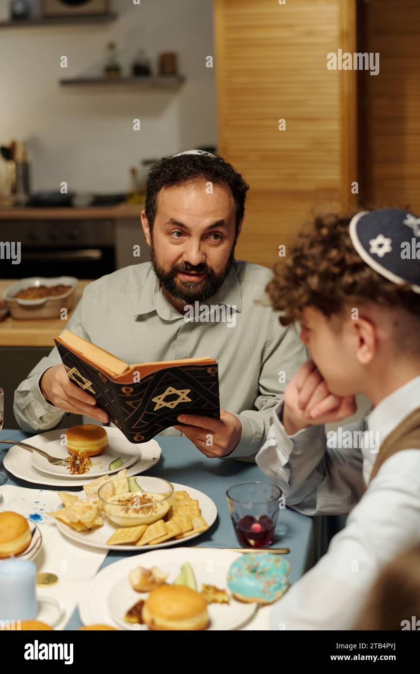 Focus on bearded mature man in kippah looking at his pre-teen son while ...