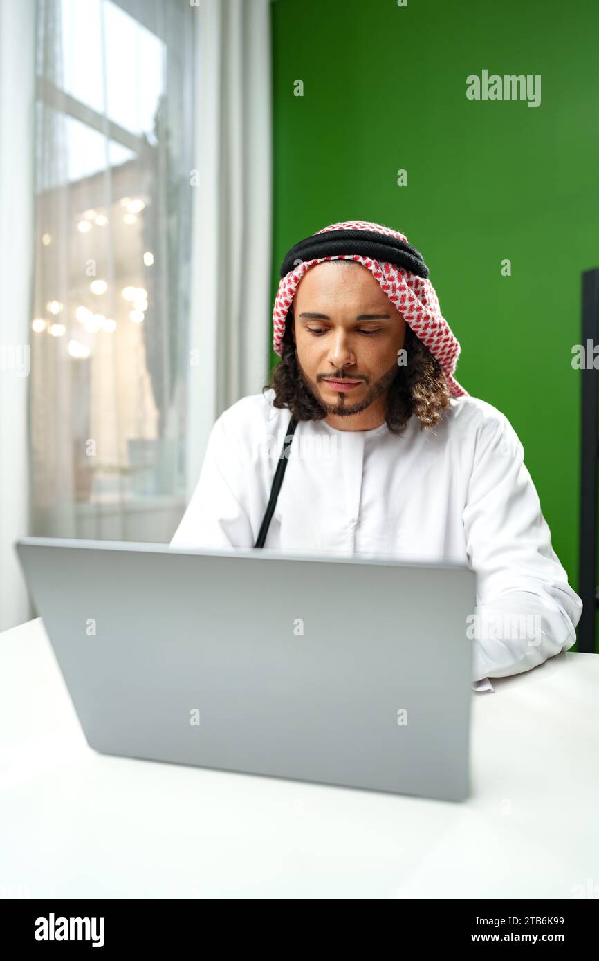 Young Arab man in traditional dress thobe working on computer in office ...