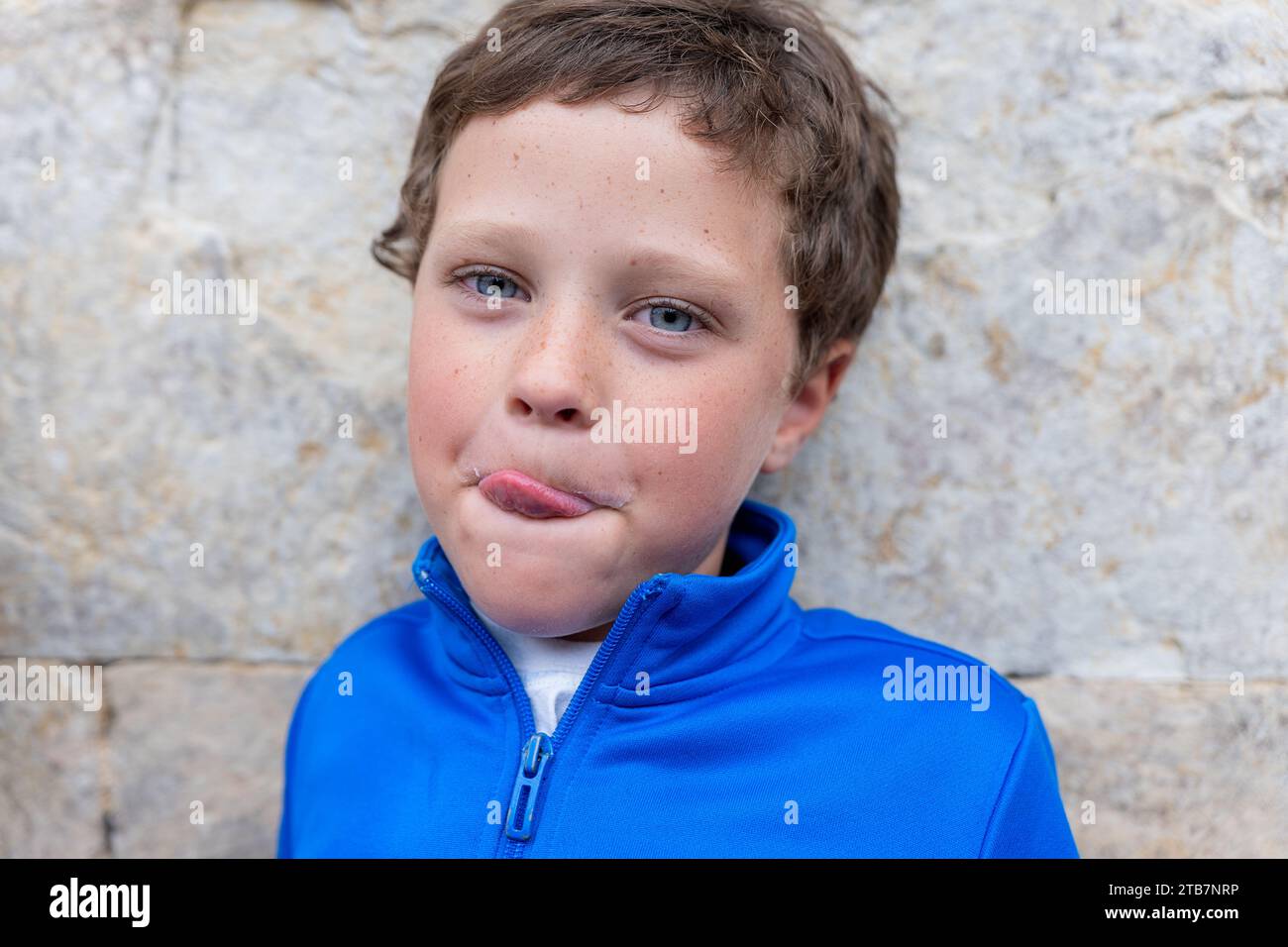A boy in a blue jacket making a funny face against a stone wall ...