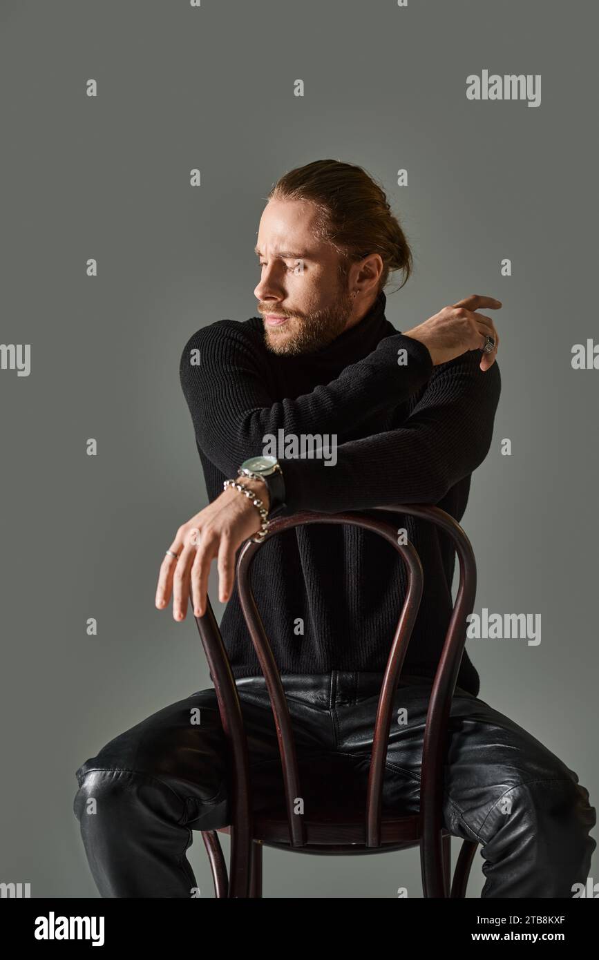 bearded man in turtleneck and black leather pants sitting on wooden chair on grey background ...