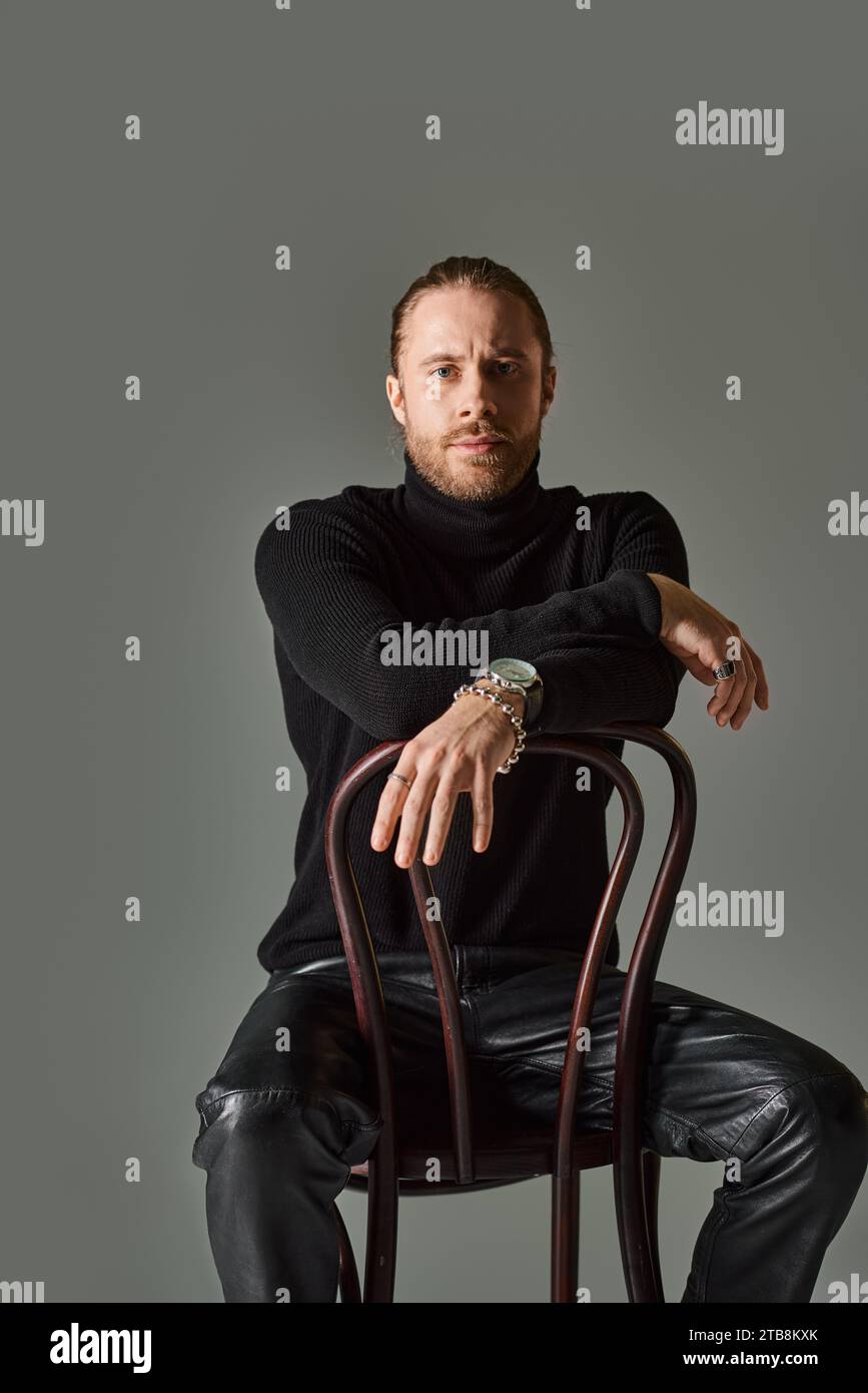 bearded man in turtleneck and black leather pants sitting on wooden chair on grey background ...