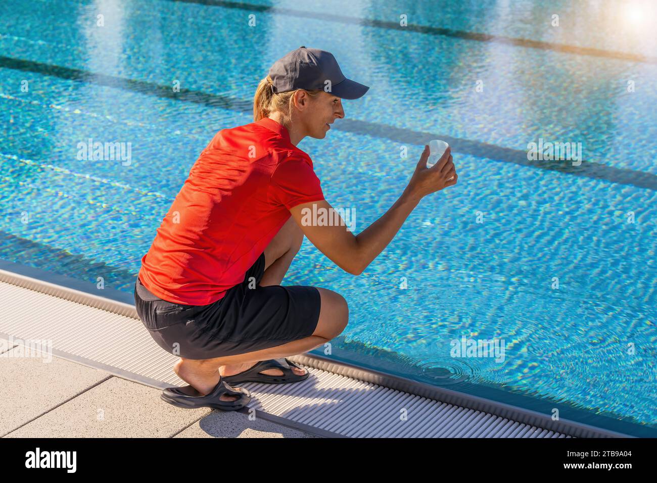 Woman squatting by a pool holding a clear container to examine water ...