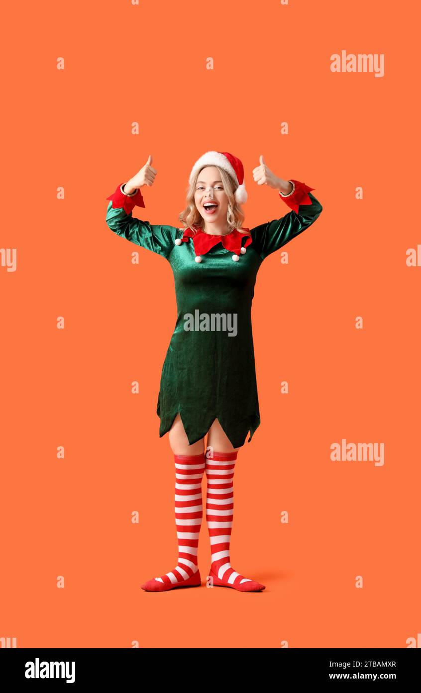 Happy young woman dressed as elf showing thumbs-up on orange background ...