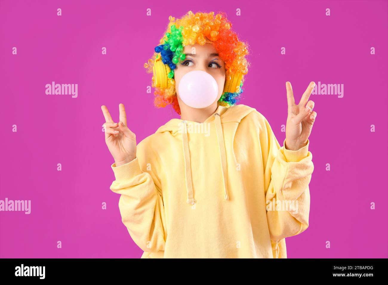 Trendy girl in clown costume blowing bubble gum while showing victory ...