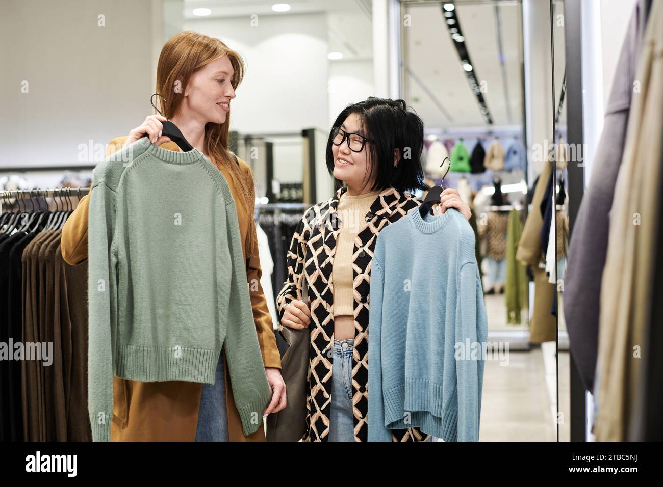 Medium long shot of two women showing clothes to each other while shopping at store Stock Photo ...