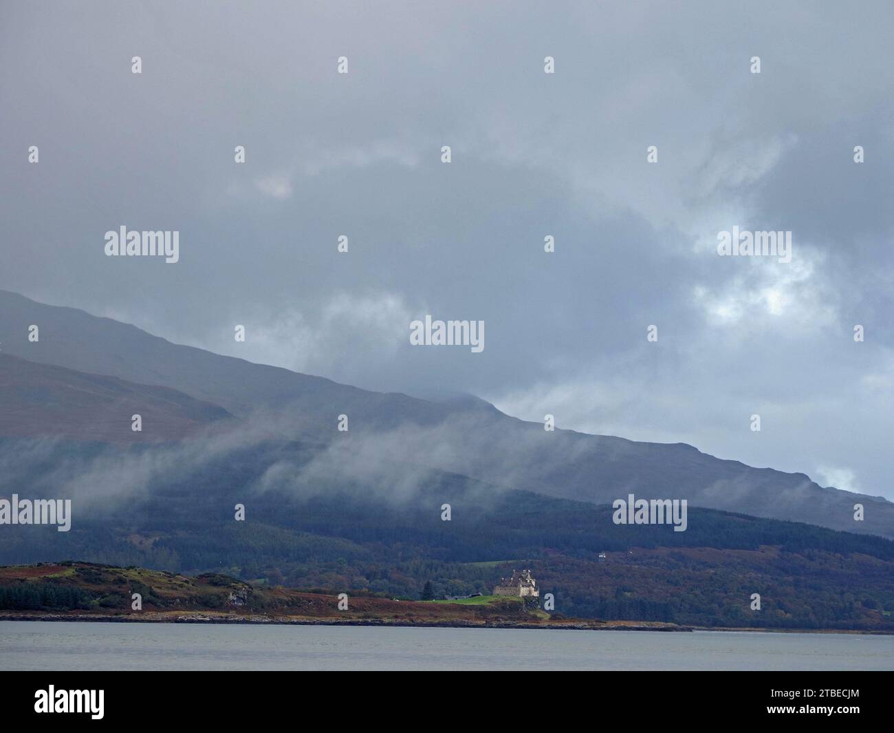 wispy clouds over wooded hillside on shoreline with sunlit stately home ...