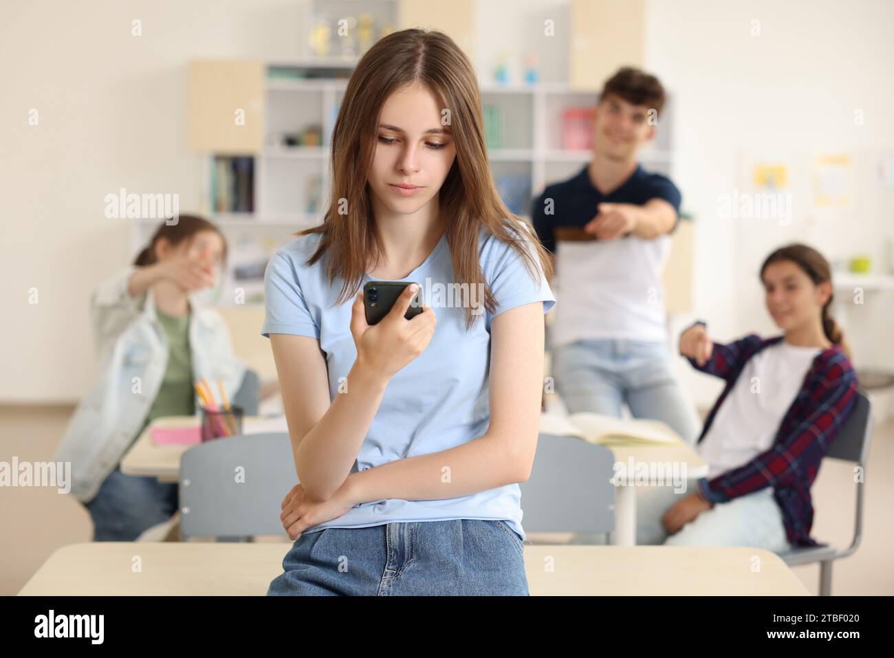 Teen problems. Lonely girl with smartphone standing separately from other students in classroom ...