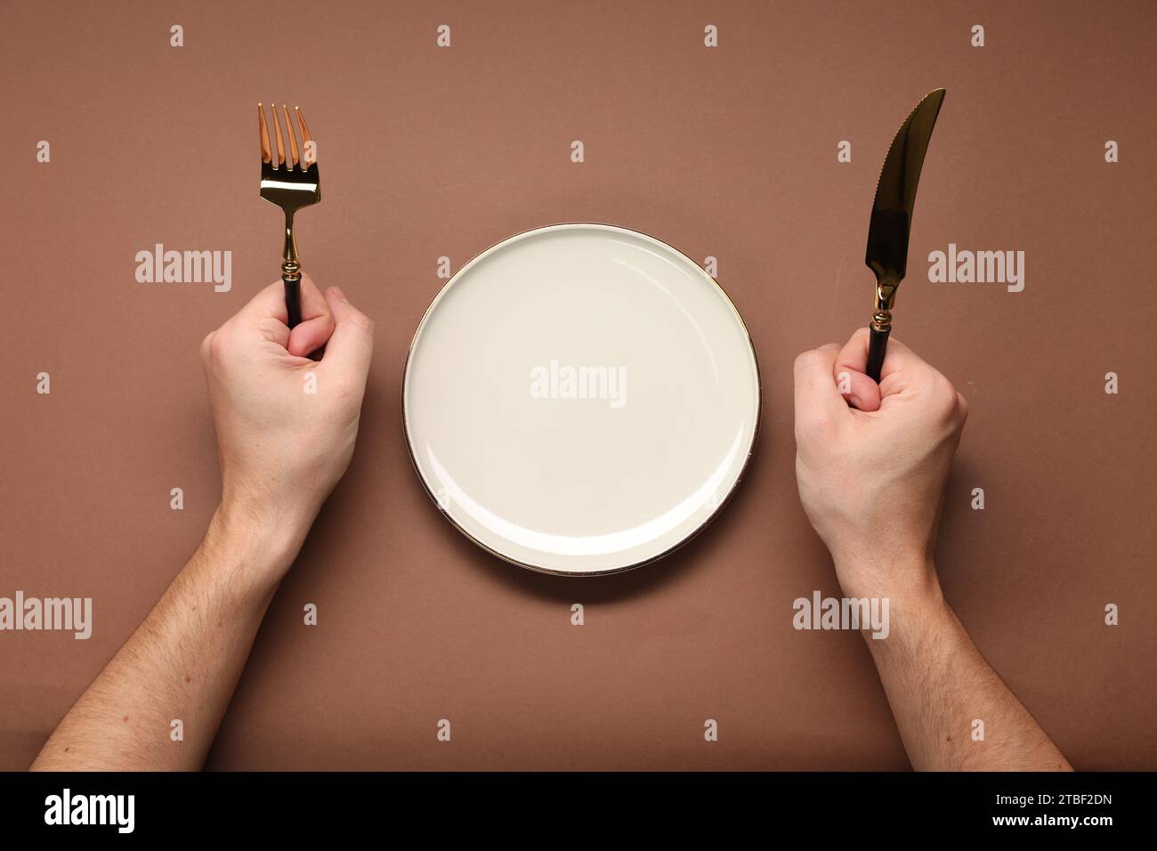 Man holding fork and knife near empty plate at brown table, top view ...