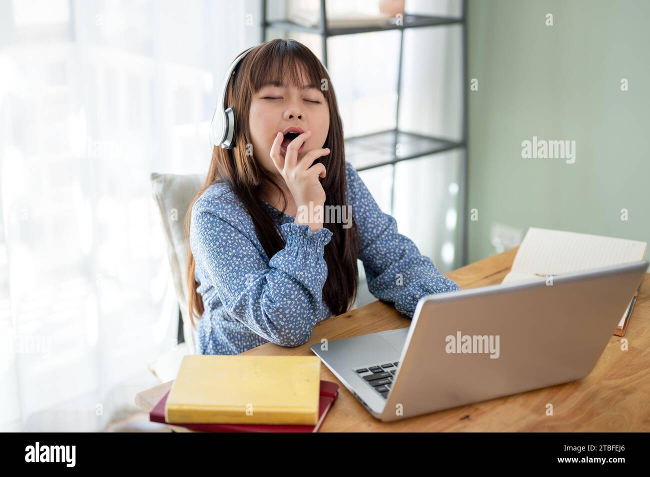 A cute young Asian girl is feeling tired and sleepy while studying online on her laptop computer ...