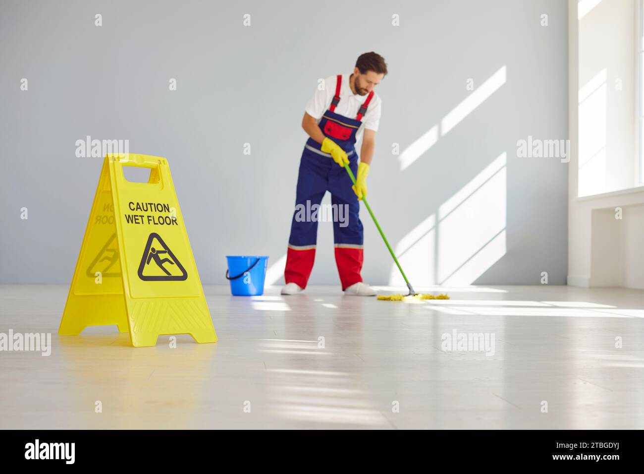 Caution wet floor sign with male young cleaner man mopping empty room ...