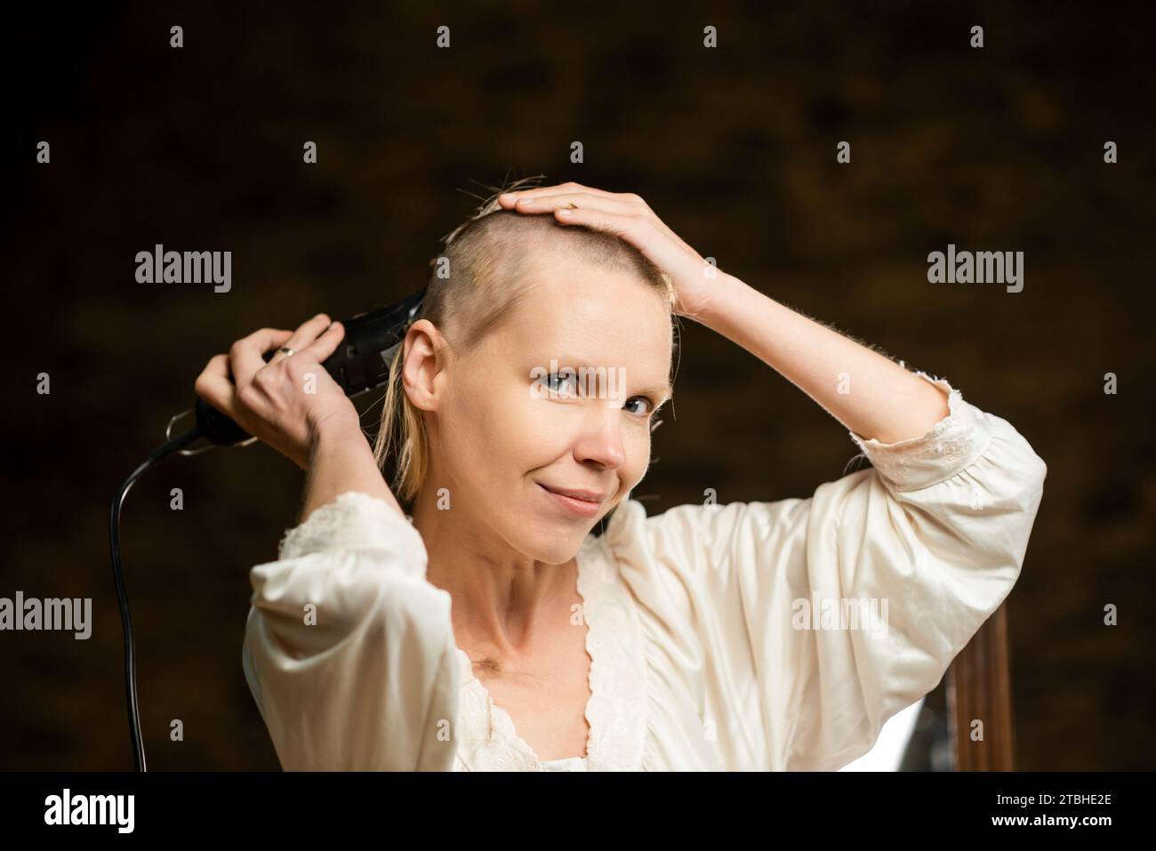 A woman smiles and cuts her hair on her head with a clipper on a dark ...