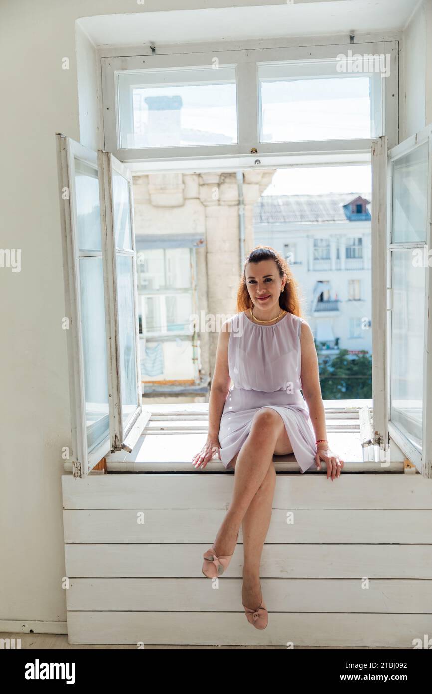 woman in summer light dress sitting on the window looking at the street ...