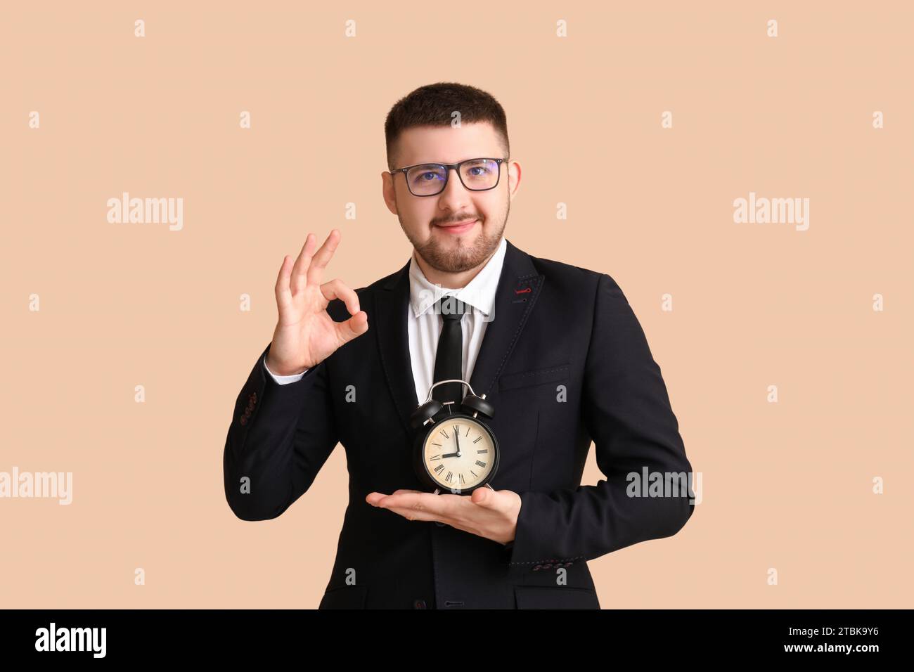 Young happy businessman with alarm clock showing OK gesture on beige ...