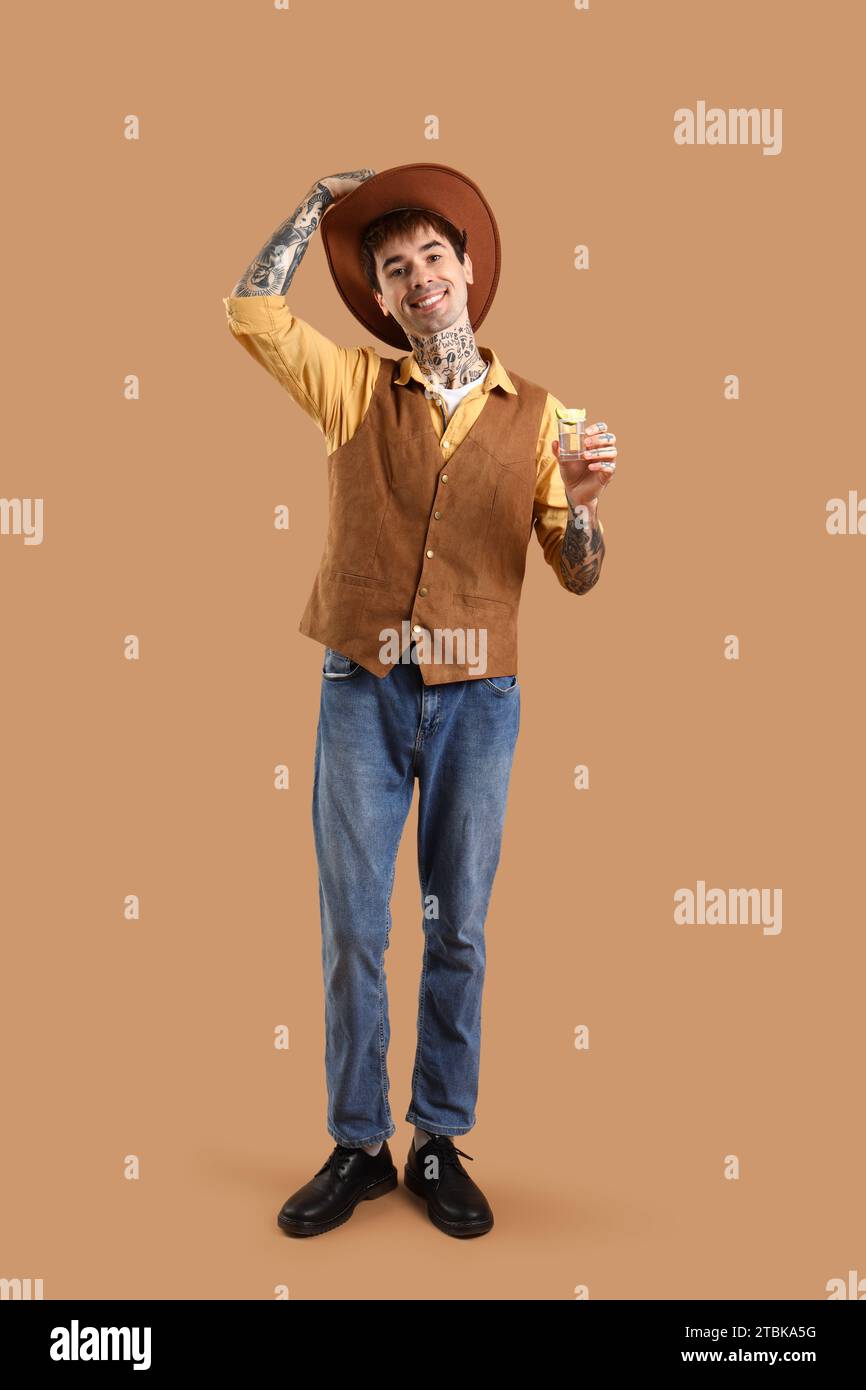 Young male bartender dressed as cowboy with shot of tequila on beige ...