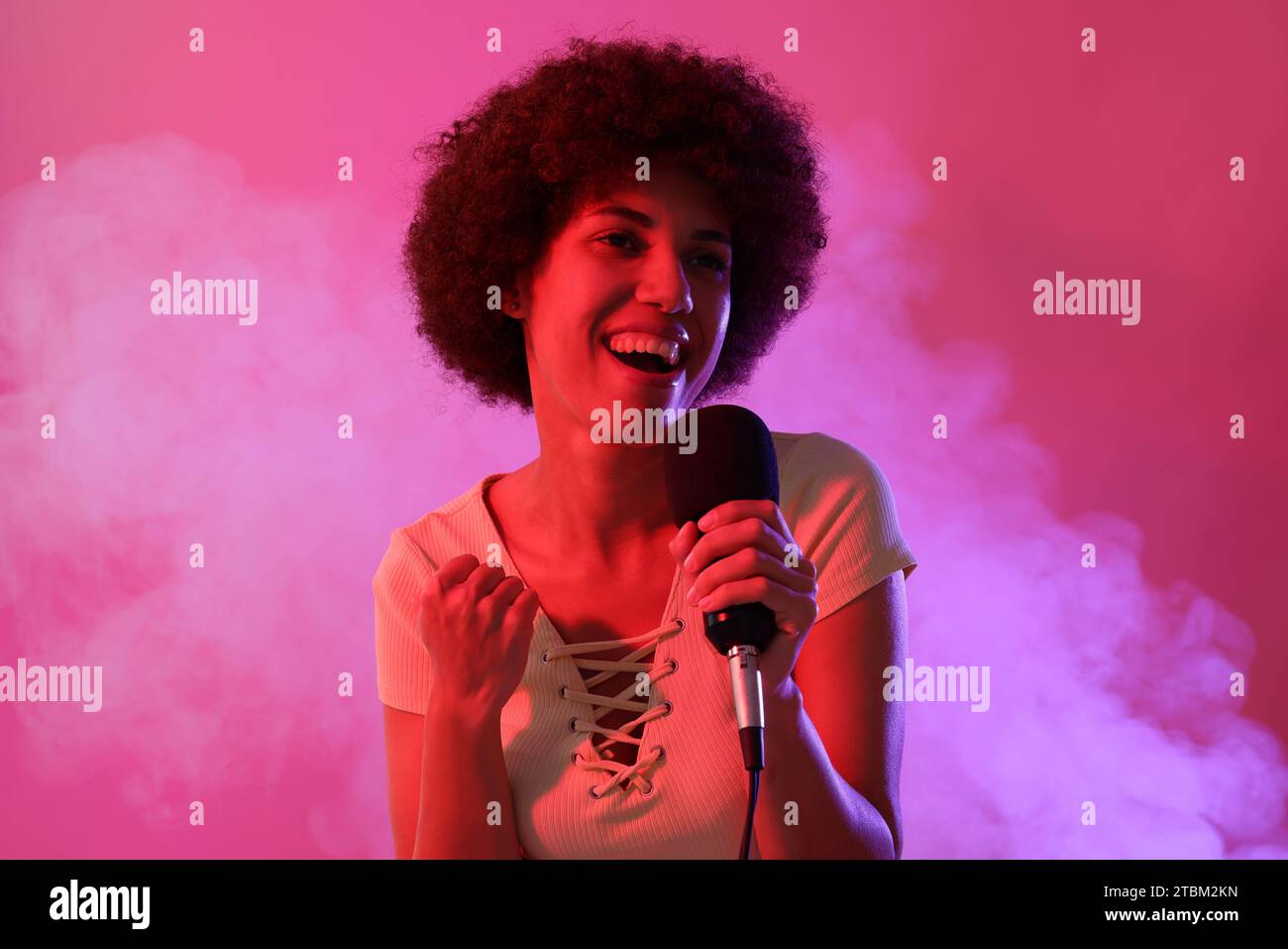 Curly young woman with microphone singing on pink background. Color ...