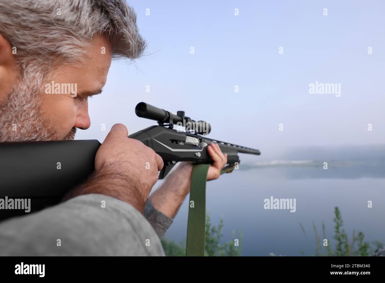 Man aiming with hunting rifle near lake outdoors, closeup. Space for ...