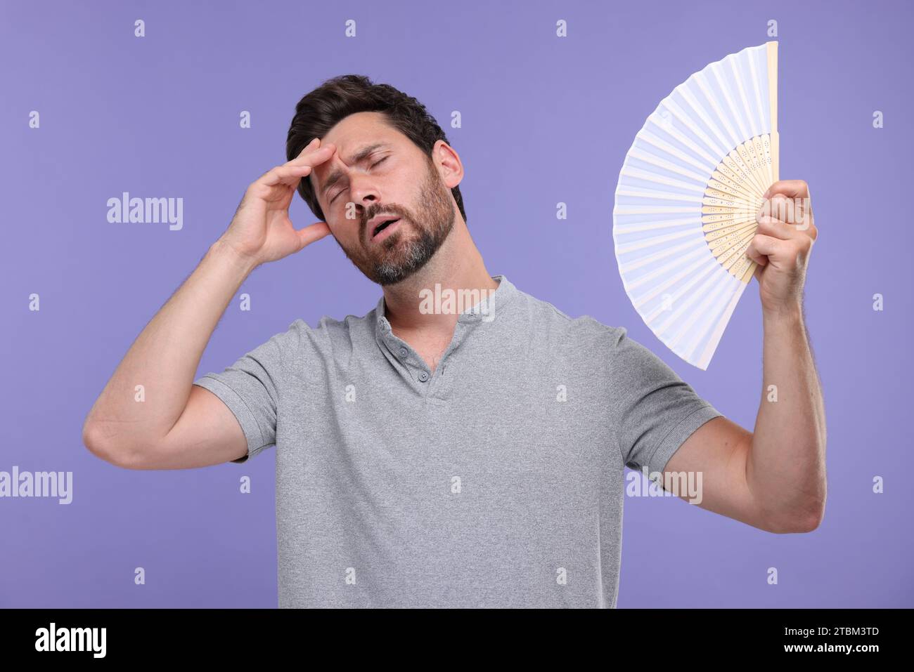 Unhappy man with hand fan suffering from heat on purple background ...