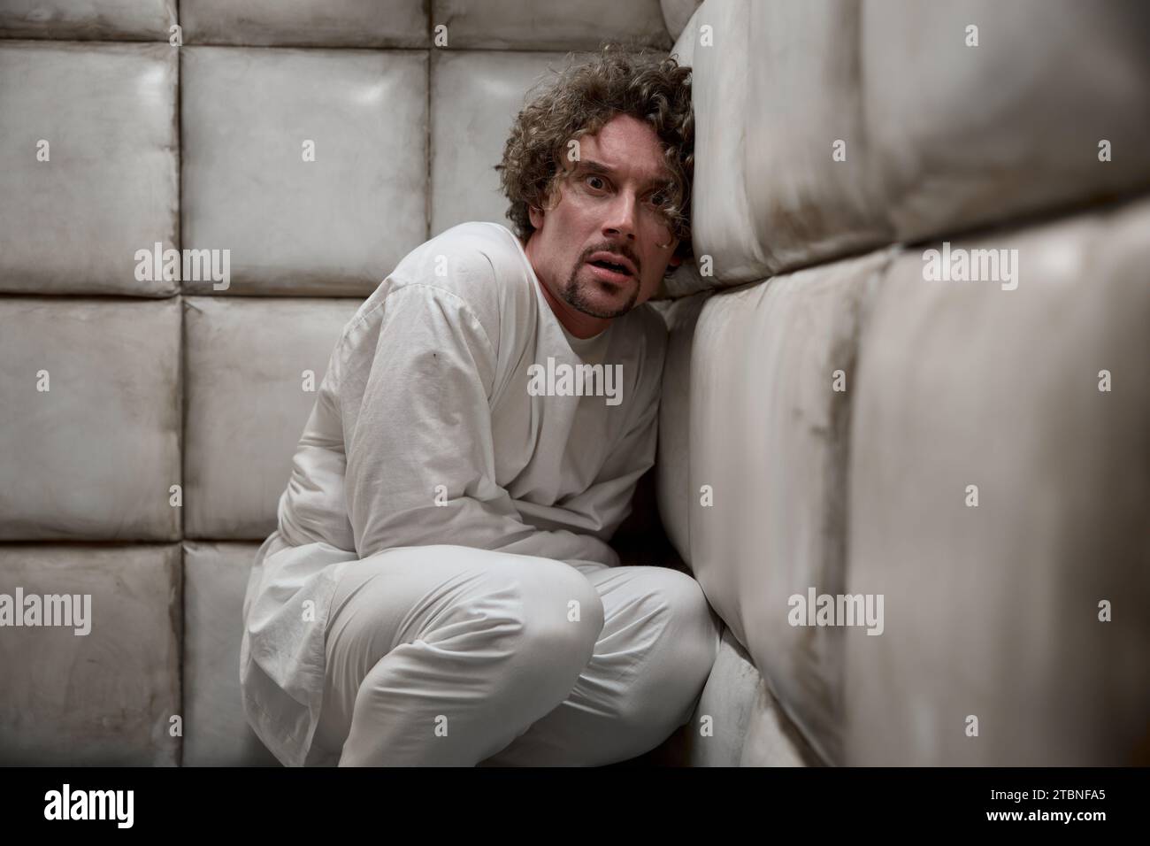 Mad scared man patient wearing straitjacket in corner of padded room ...