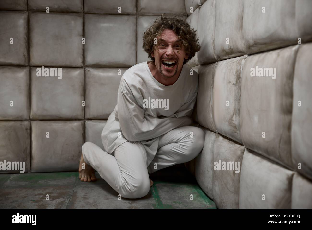 Mad crazy man patient wearing straitjacket laughing terrifying sitting in corner of padded room ...