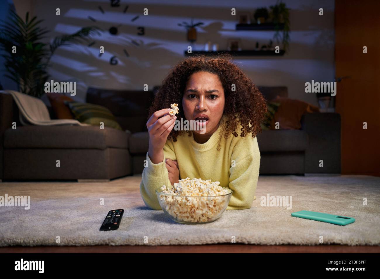 Portrait latina surprised afro woman lying on living room carpet eating ...