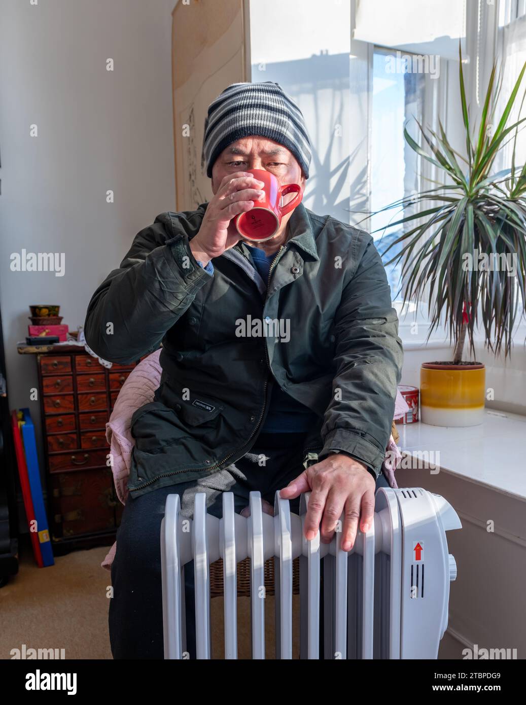 An elderly man in the living room sitting by a portable electric heater ...