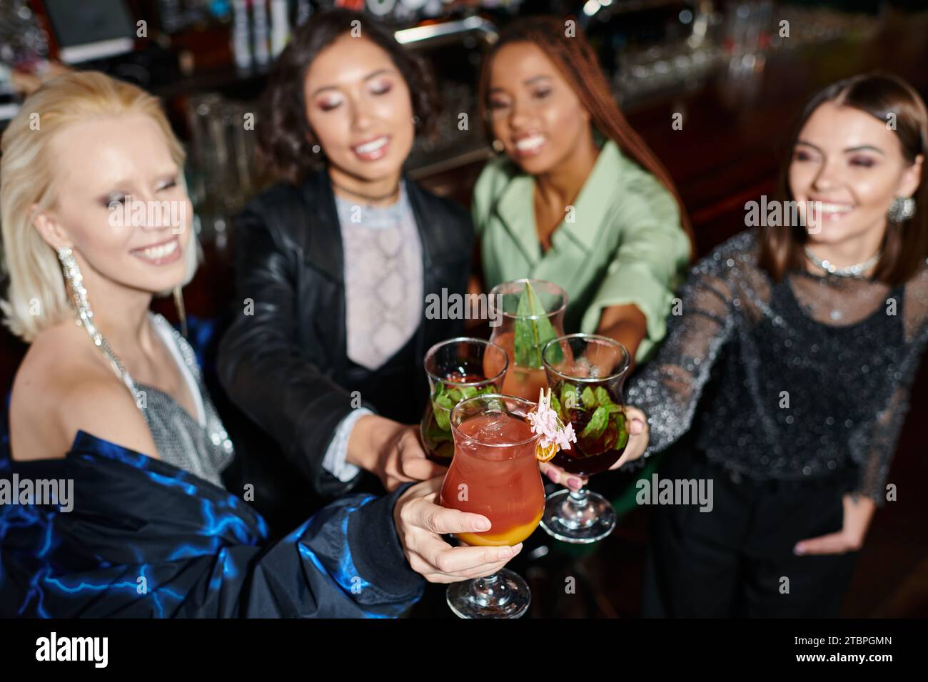 smiling and elegant multiethnic girlfriends clinking cocktails in bar ...
