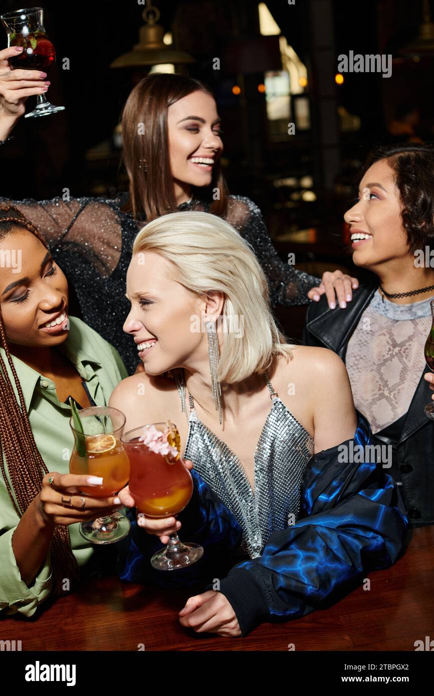 happy alluring women clinking cocktail glasses near multiethnic ...