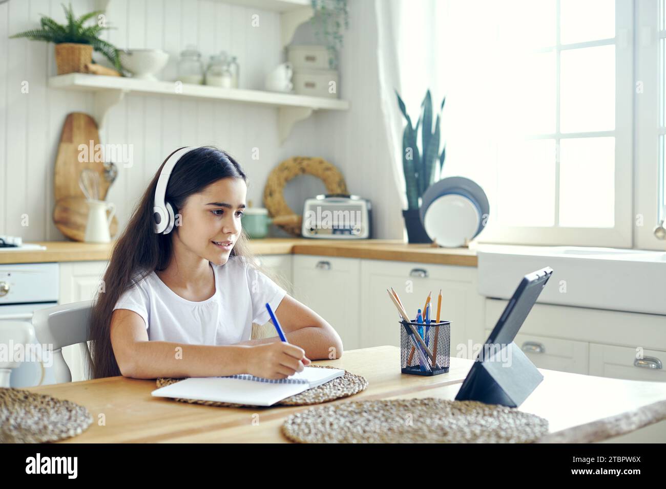 Pretty pre-teen 12s girl in wireless headphones sit at table e-learning ...