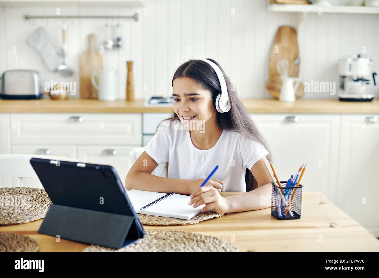 Pretty pre-teen 12s girl in wireless headphones sit at table e-learning ...
