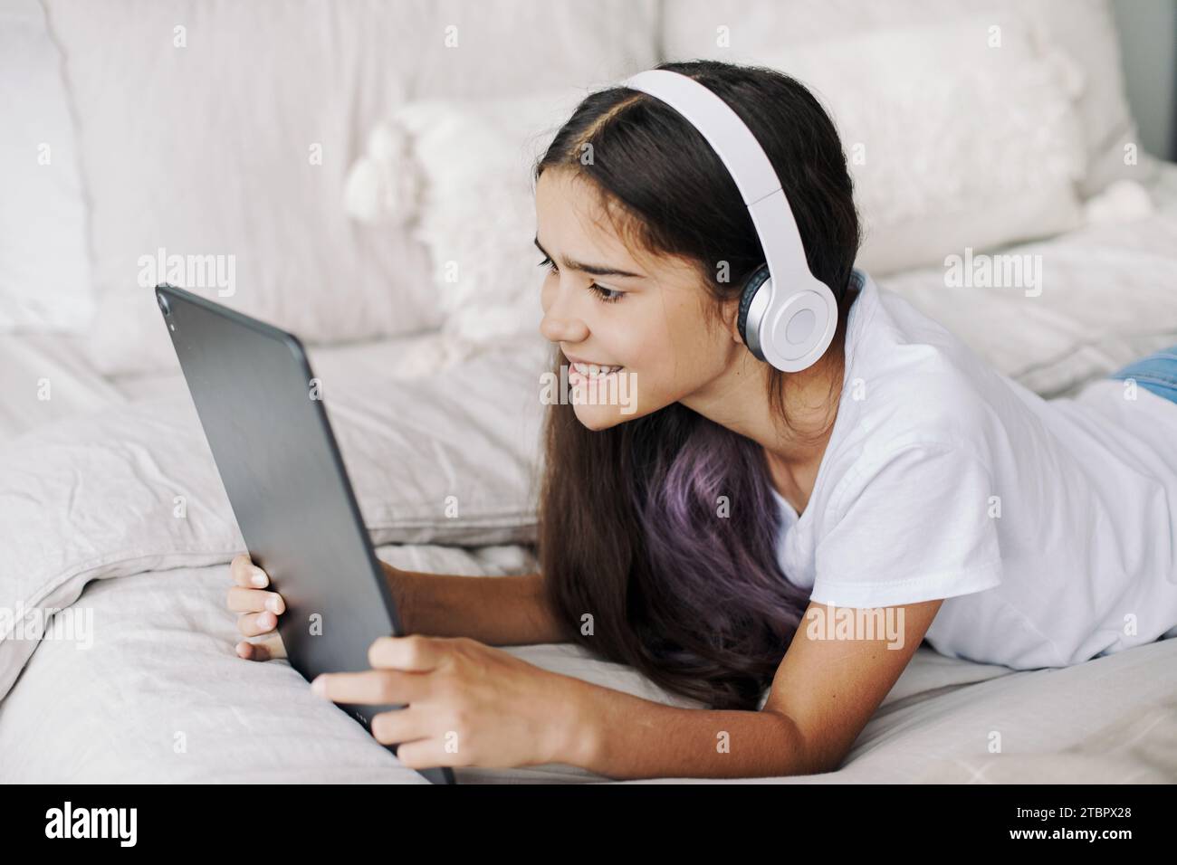 Adorable pre-teen girl in wireless headphones lying on bed with digital tablet device. Young ...